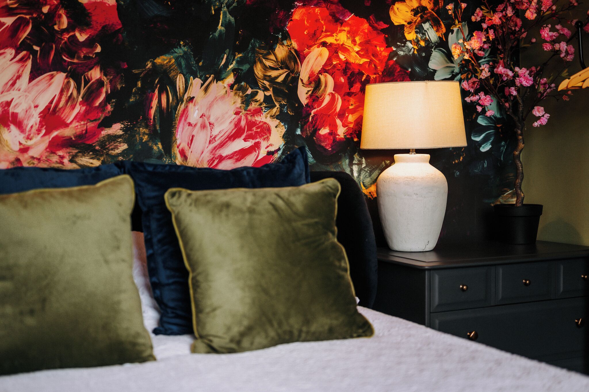 Quarto com cama, almofadas verdes, mesa de cabeceira, abajur branco e um mural floral na parede.
