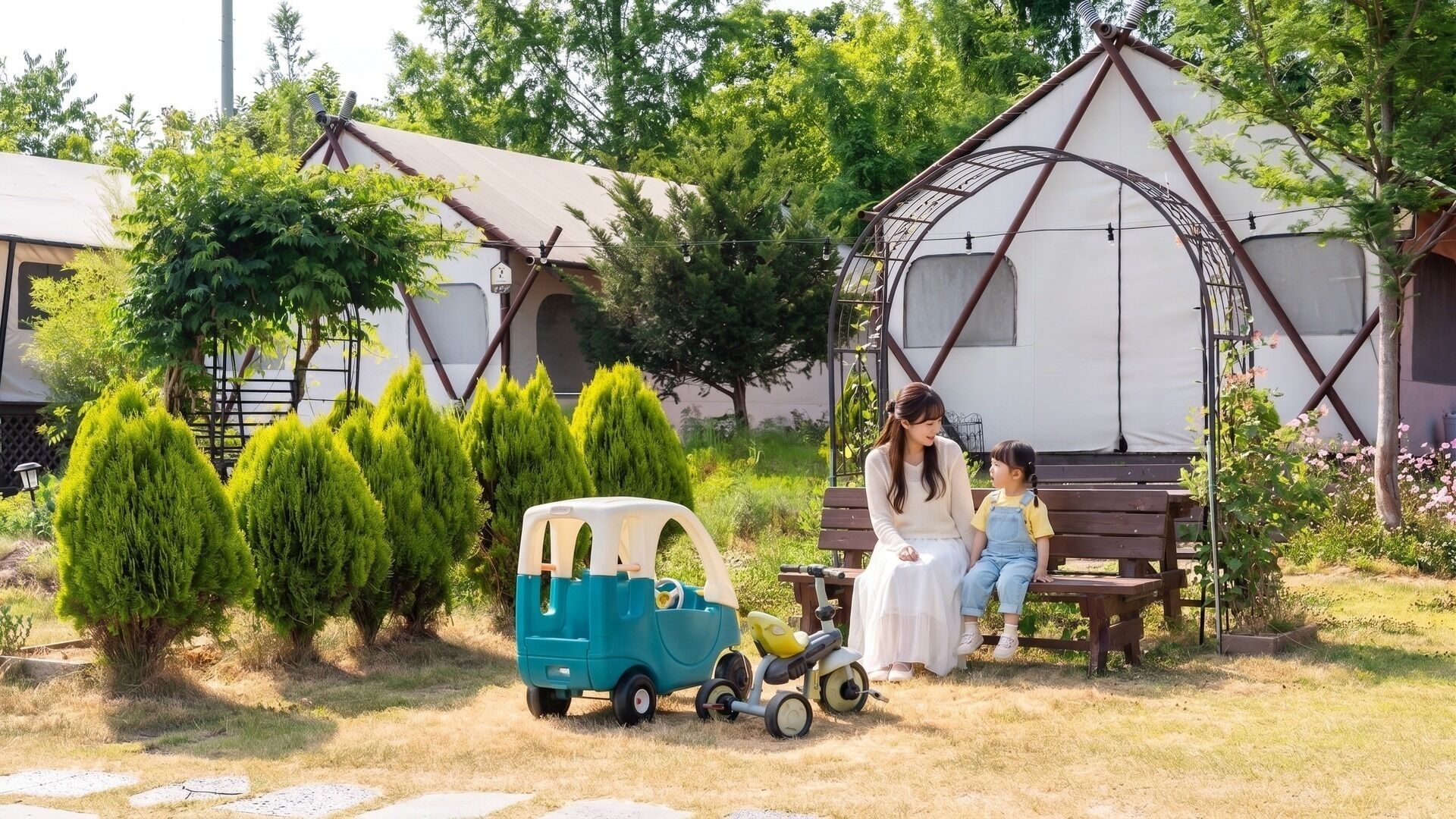 Um camping com tendas, um pequeno carro de brinquedo azul e uma mulher e criança sentadas em um banco.