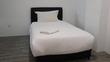Uma cama de solteiro com colchão branco e cabeceira preta em quarto com piso de madeira e paredes brancas.