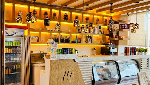Interior de café moderno com vigas de madeira, luminárias pendentes e um balcão central com o desenho de uma xícara de café.
