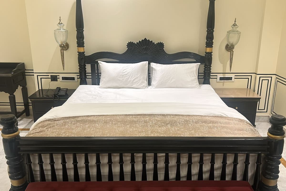 Uma cama de dossel com lençóis brancos e um cobertor dourado, ladeada por mesas de cabeceira com abajures, em um quarto com paredes bege e detalhes em preto.