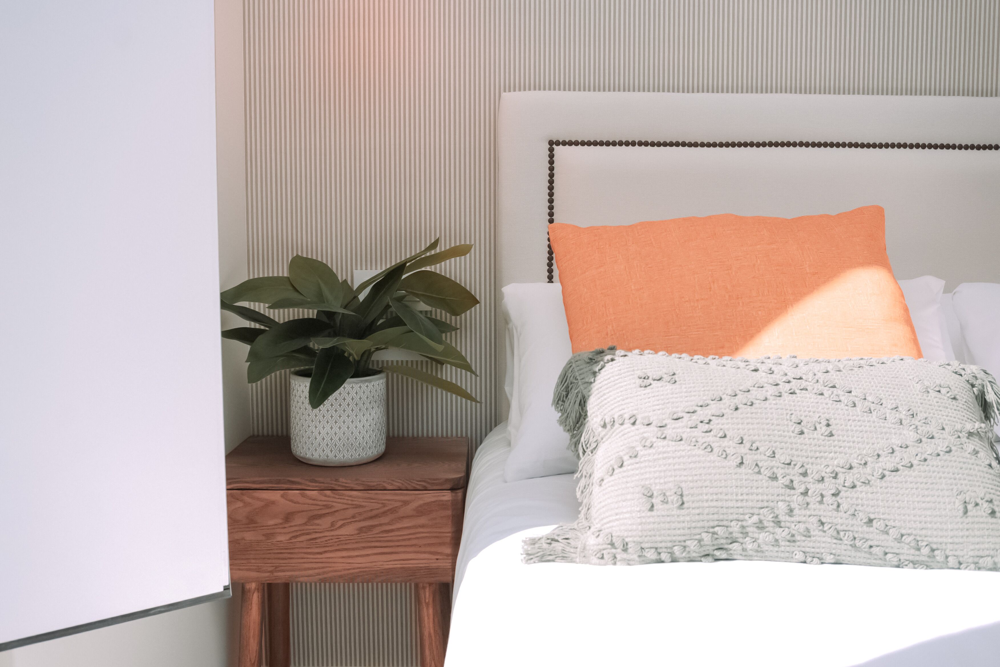 Cama bem arrumada com cabeceira branca, travesseiro laranja e cobertor branco. Ao lado da cama, um criado-mudo de madeira com uma planta em vaso.