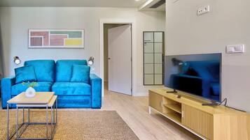 Uma sala de estar com um sofá azul, uma mesa de centro, uma TV em um suporte de madeira e um quadro na parede.