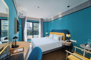 Quarto de hotel com cama, mesa de cabeceira, abajur, escrivaninha com computador, cadeira e vista da cidade através de amplas janelas.