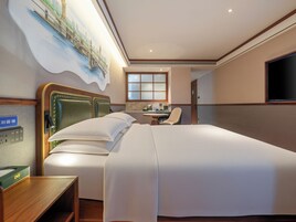 Quarto de hotel com cama grande, escrivaninha, cadeira, televisão e um quadro na parede.