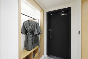 Um corredor com uma porta preta, uma prateleira de madeira com chinelos e um suporte com roupões cinzas.