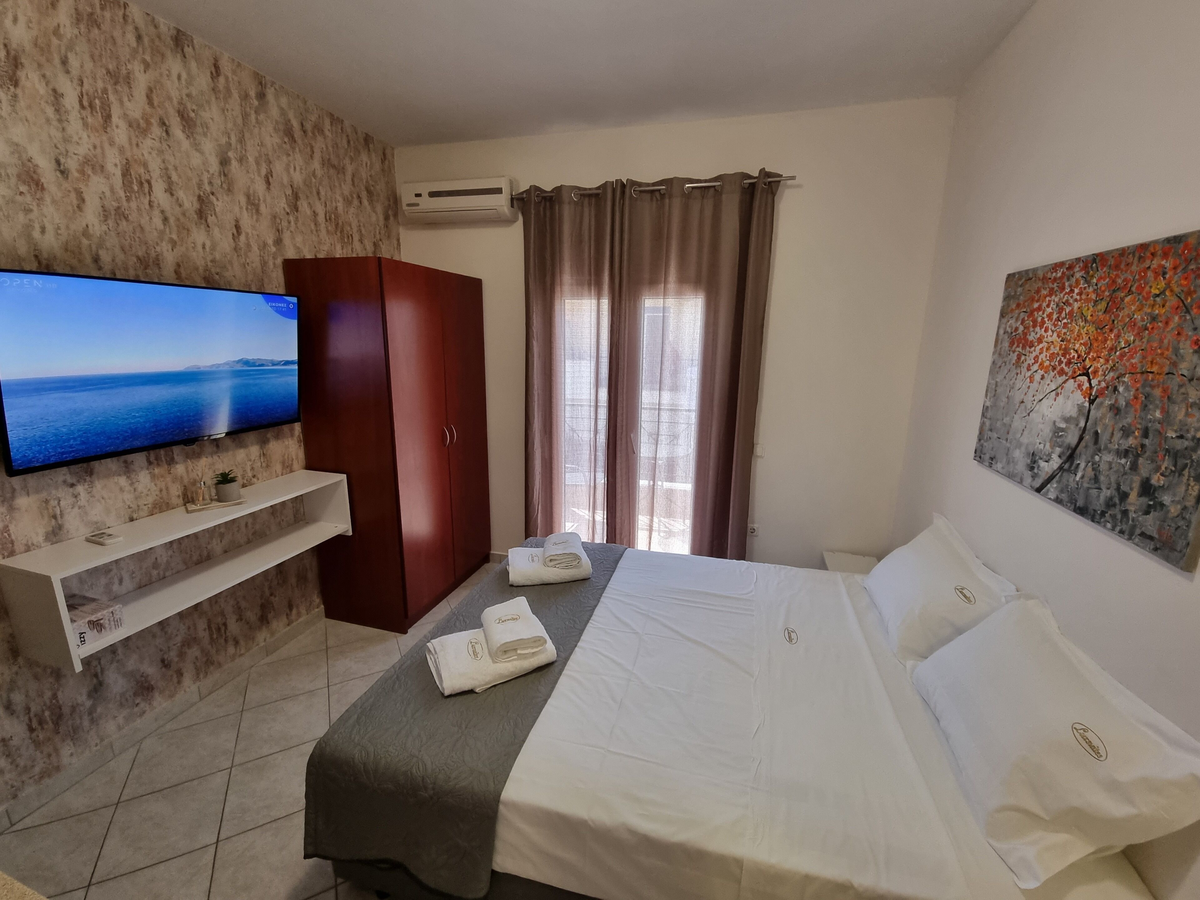 Quarto de hotel com cama, televisão, guarda-roupa, um quadro e janela com cortinas.