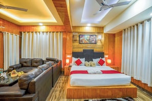 Quarto de hotel com decoração em madeira, uma cama grande com roupa de cama vermelha e branca, um sofá de couro e um ventilador de teto.