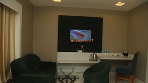 Quarto moderno com TV de tela plana na parede, mesa com tampo de vidro, duas poltronas verdes e uma cadeira de madeira.
