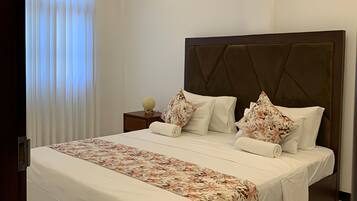 Quarto de hotel com cama, ventoinha no teto, mesa de cabeceira, quadro pendurado na parede e janela com cortinas.