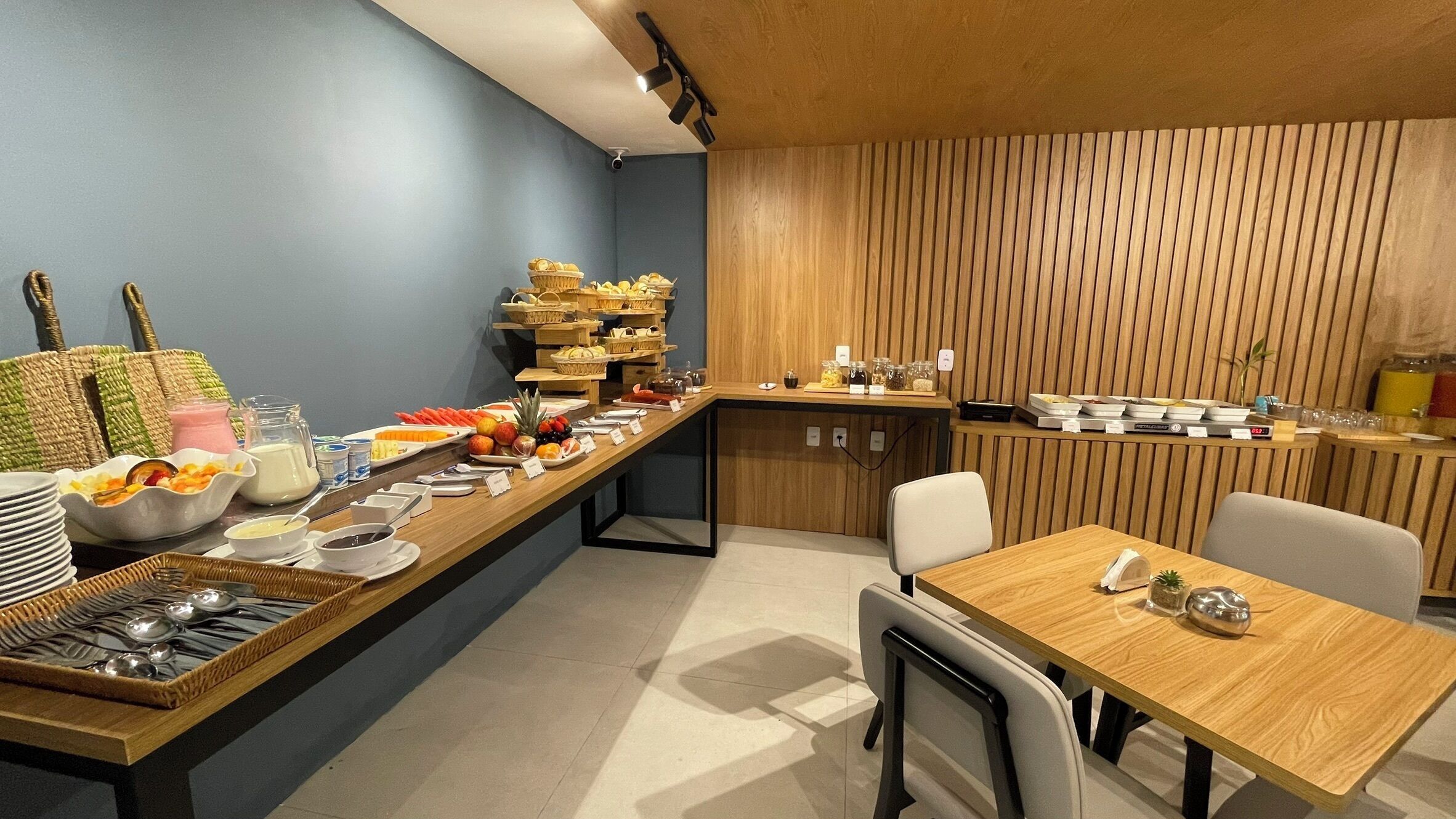 Buffet de café da manhã com diversos alimentos e área de estar com cadeiras e mesa.