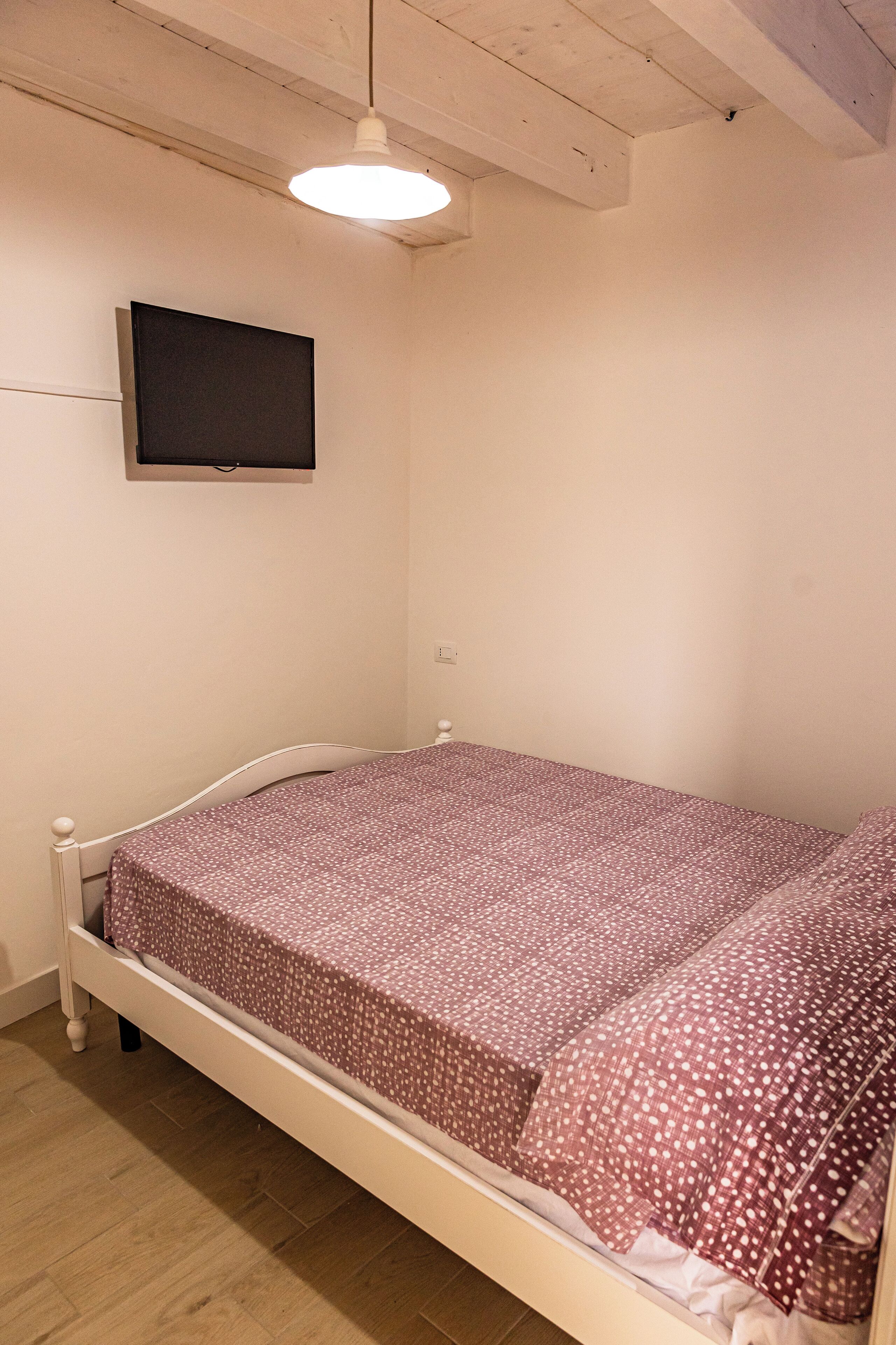 Uma cama de solteiro com estrutura branca e colcha vermelha de bolinhas em quarto com piso de madeira e teto branco.