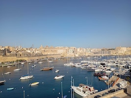 Uma marina com vários barcos atracados, céu claro e paisagem urbana com edifícios históricos ao fundo.