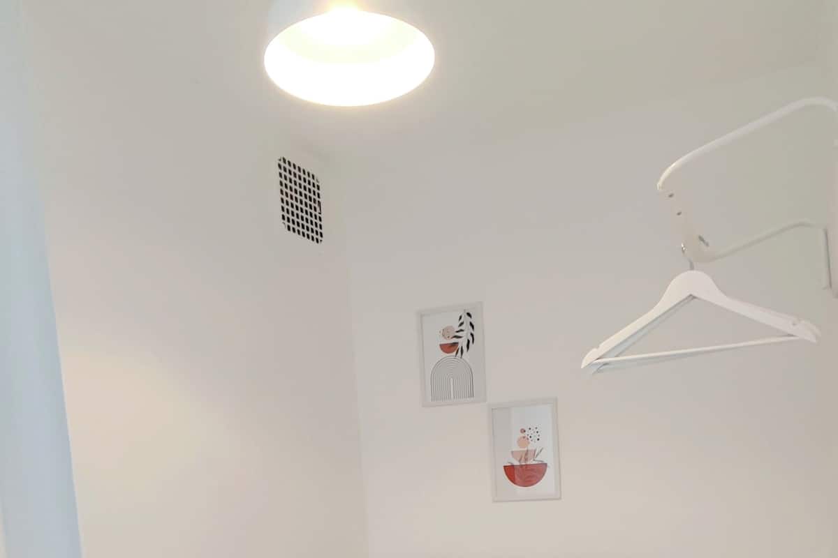 Uma cama de solteiro com cabeceira escura, roupa de cama branca, um travesseiro marrom, um cabide branco e uma pequena lixeira.