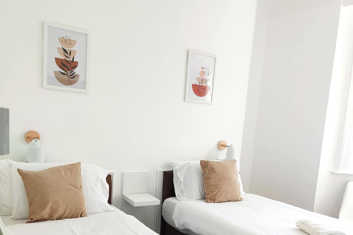Quarto de hotel com duas camas de solteiro, roupa de cama branca, uma pequena mesa de cabeceira com abajur e uma obra de arte na parede.