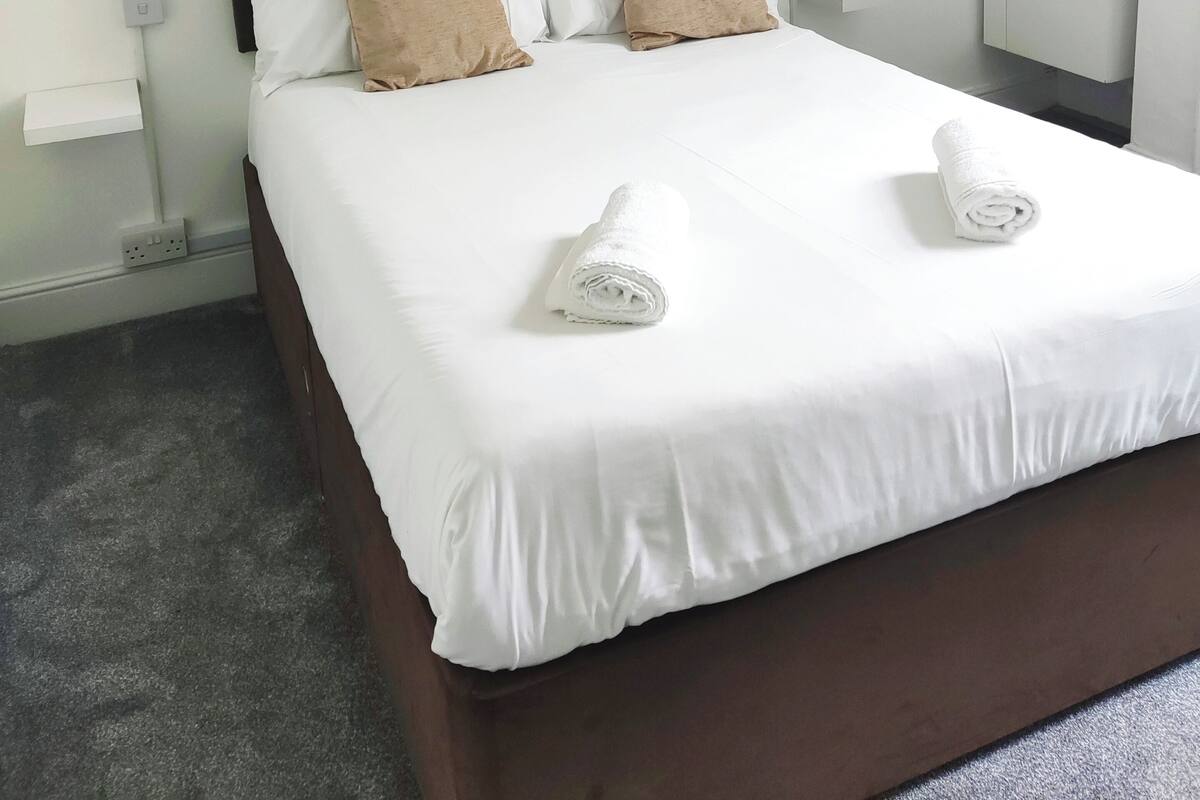 Cama bem arrumada com lençóis brancos e duas toalhas enroladas, cabeceira marrom escura e três obras de arte penduradas na parede.