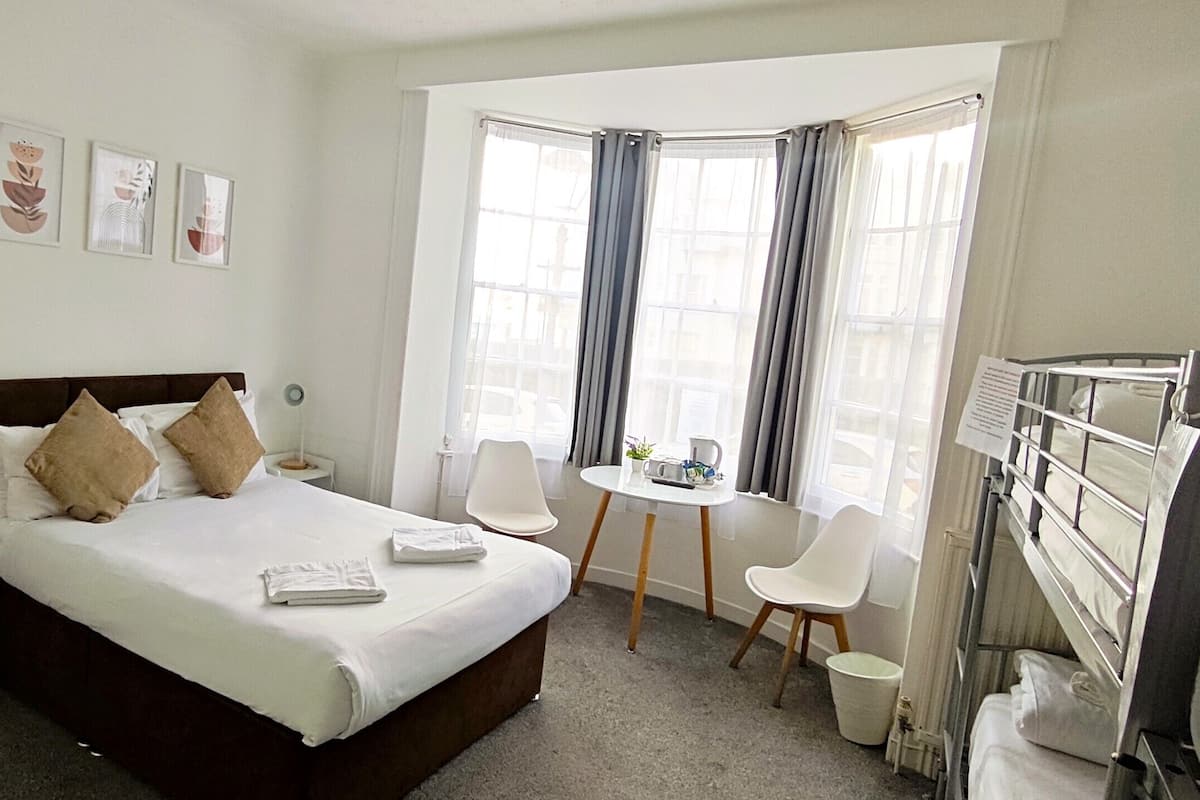 Quarto de hotel com cama, mesas de cabeceira, escrivaninha, cadeiras, janela com cortinas e uma cama beliche.