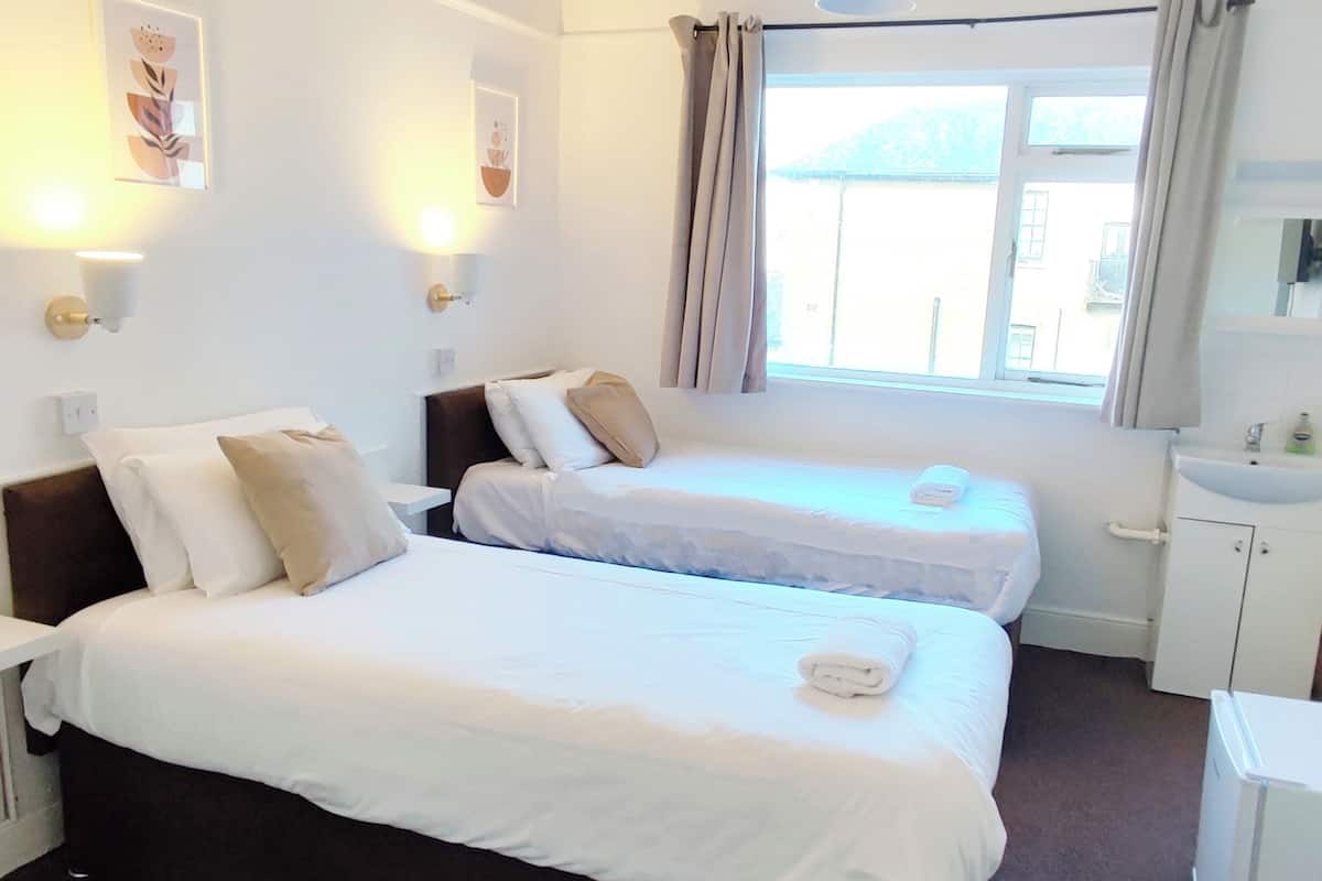 Quarto de hotel com duas camas, uma pequena mesa, uma janela com cortinas e luminárias fixadas na parede.