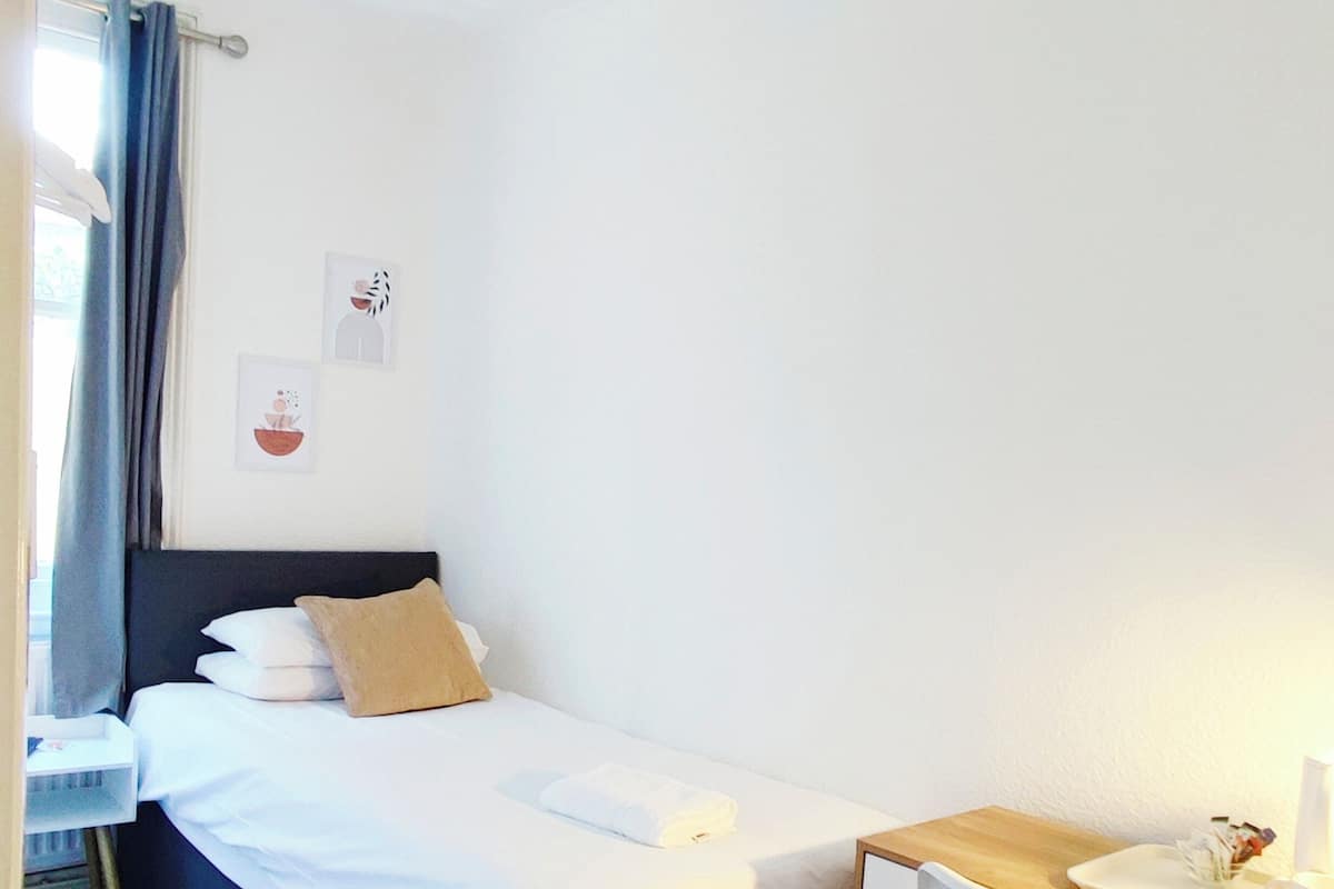 Quarto de hotel com cama, escrivaninha, cadeira, luminária pendente e janela com cortinas.