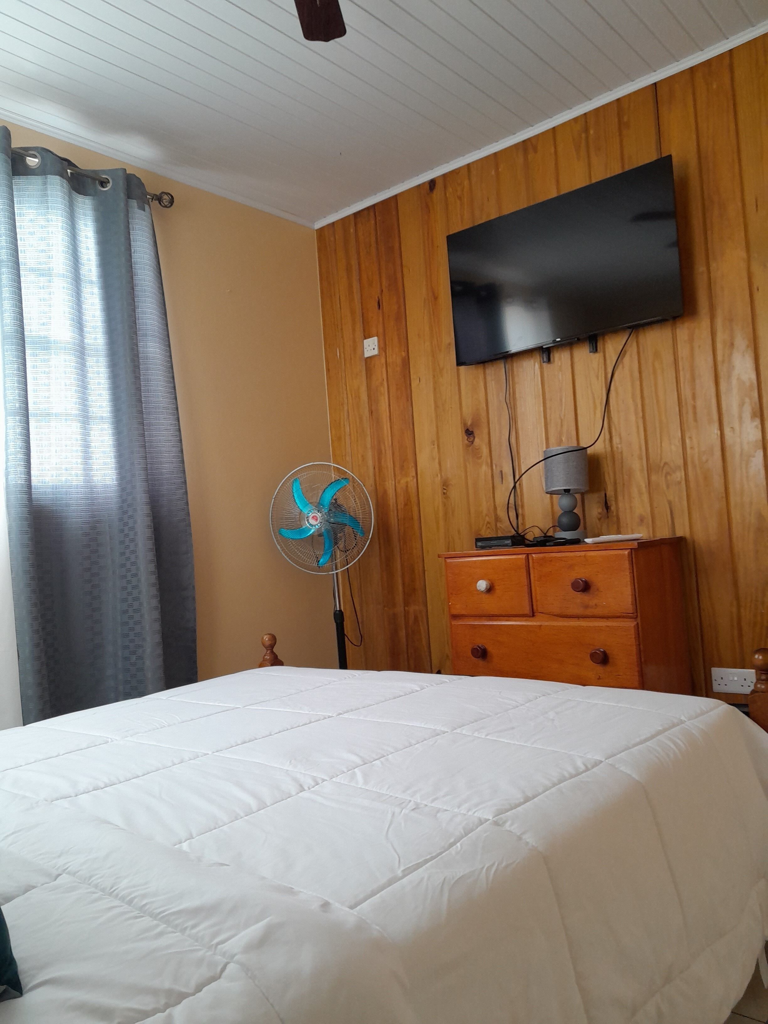 Quarto com cama, cómoda de madeira, televisão na parede, ventoinha no teto e janela com cortinas.