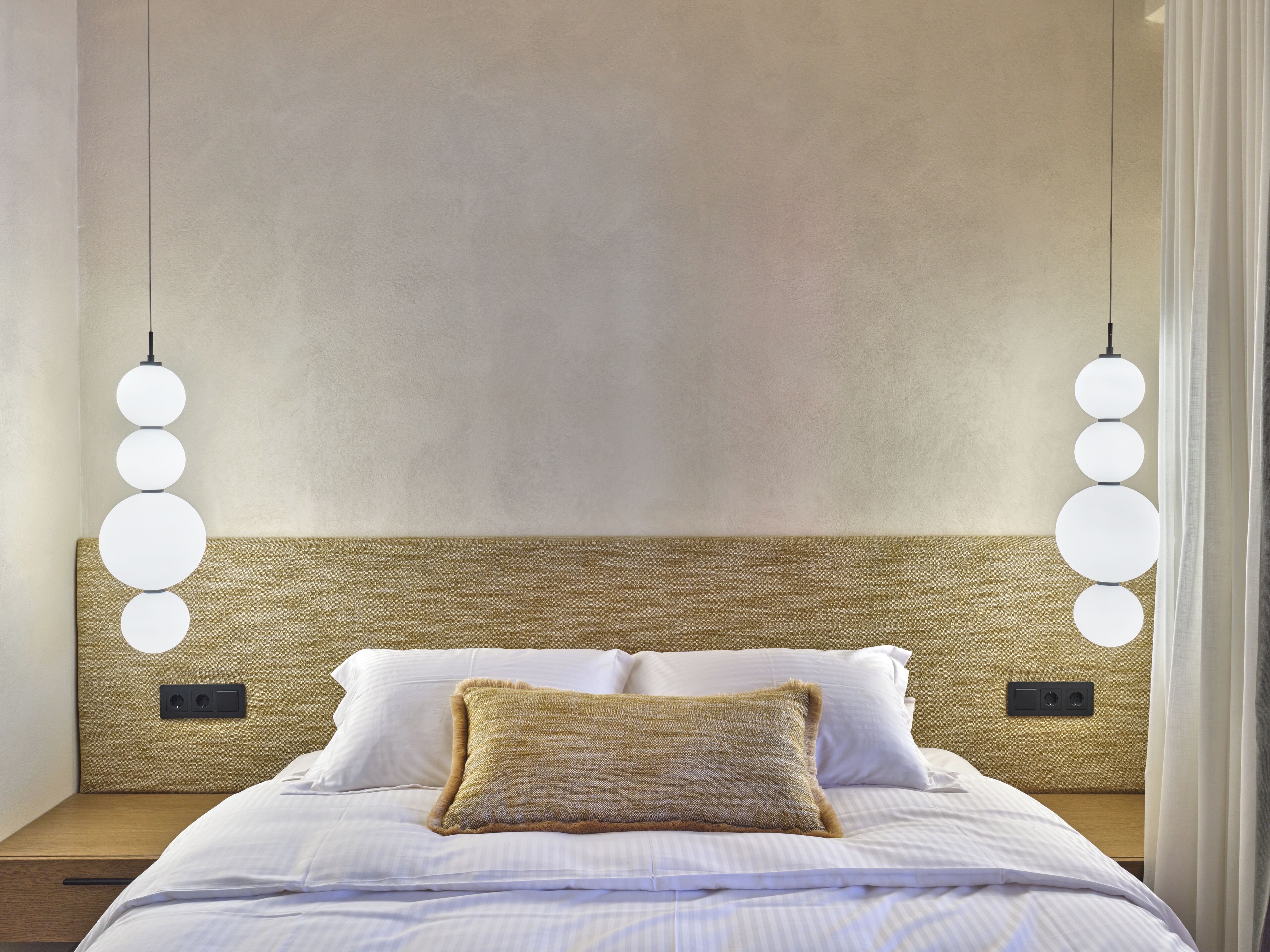 Cama bem arrumada com cabeceira de madeira, duas mesas de cabeceira com abajures e uma parede lisa.