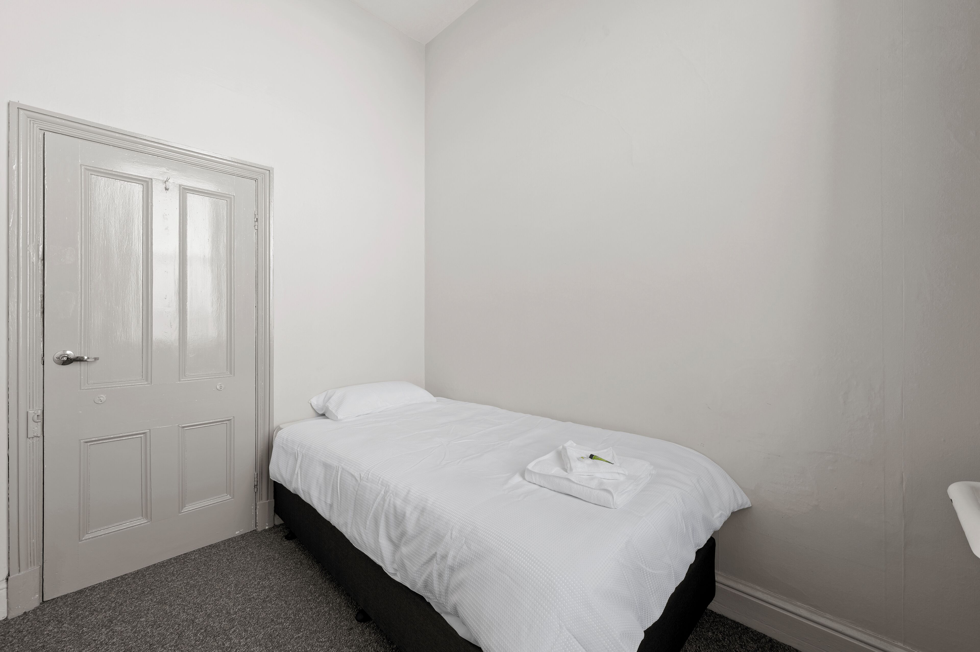 Uma cama de solteiro num quarto com paredes brancas e porta fechada.