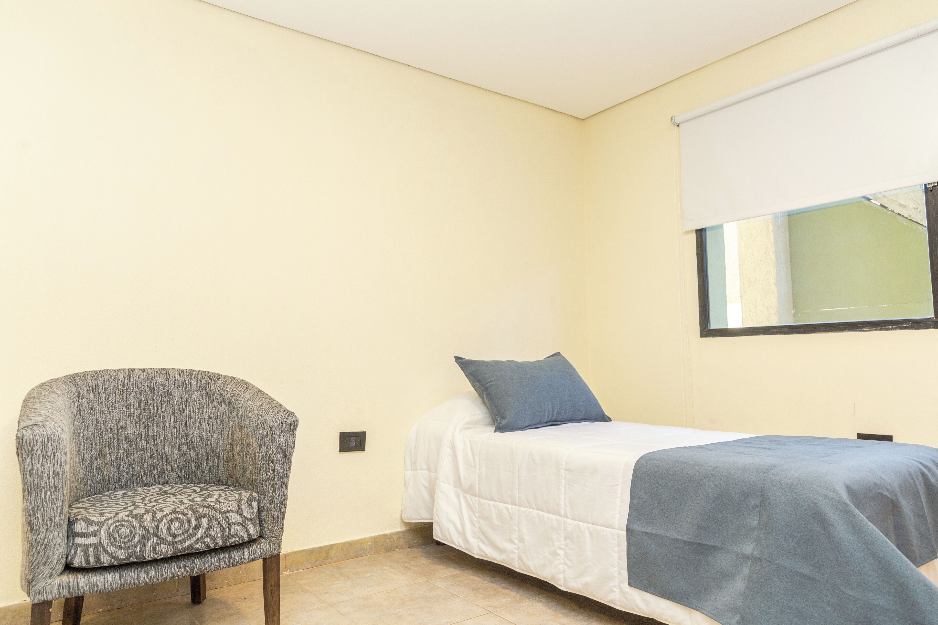 Quarto com uma cama de solteiro, uma cadeira e uma janela com cortina.