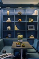Quarto com sofá azul, mesa com vaso e frutas, e estante com diversos objetos.