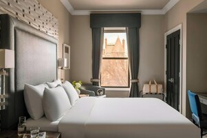 Quarto de hotel com uma cama grande, uma escrivaninha, uma cadeira, uma janela com cortinas e vista para um edifício.