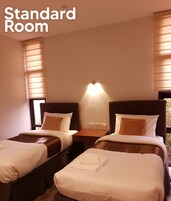 Quarto padrão com duas camas, cada uma com cabeceira, mesinhas de cabeceira e luminária fixada na parede.