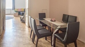 Uma sala de jantar com mesa de vidro e cadeiras pretas, um sofá ao fundo, e um chão de madeira com padrão herringbone.