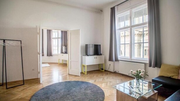 Uma sala de estar moderna com piso de madeira, uma mesa de centro de vidro, um sofá, uma TV sobre um suporte branco e uma janela grande com cortinas.