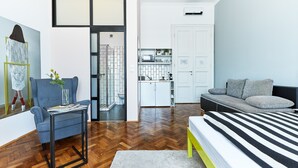 Quarto moderno com uma cama, um sofá cinza, uma mesinha de centro com um vaso e vista para um banheiro e cozinha.