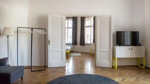 Uma sala de estar ampla com piso de madeira, um tapete cinza, uma TV em um armário branco e uma cama ao fundo.