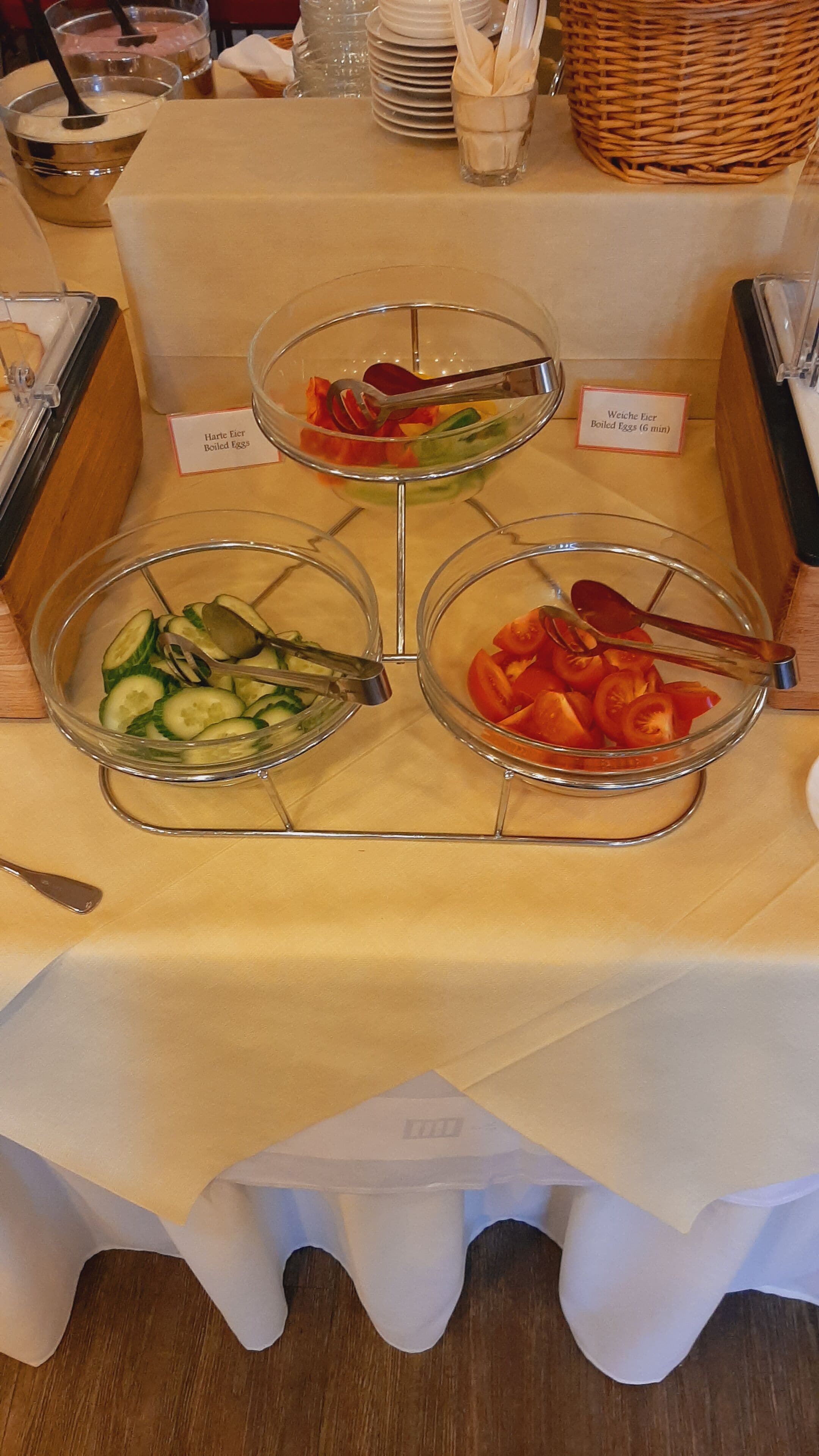 Uma mesa de buffet com três recipientes de vidro contendo pepinos fatiados, tomates e ovos cozidos.