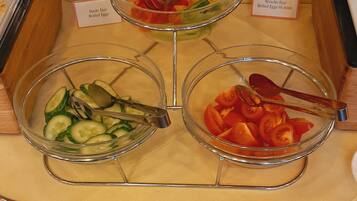Uma mesa de buffet com três recipientes de vidro contendo pepinos fatiados, tomates e ovos cozidos.