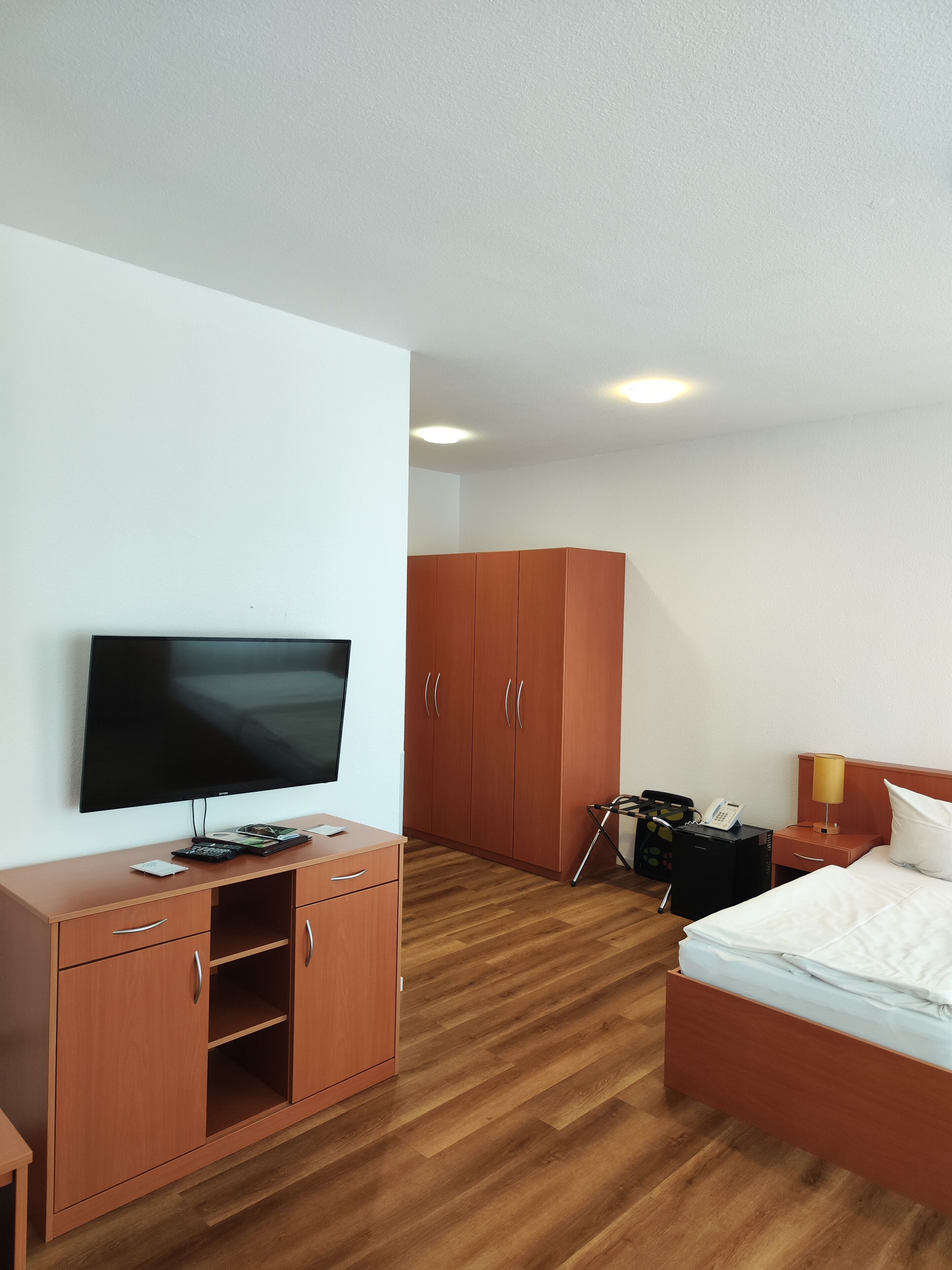 Quarto de hotel com cama, televisão, armários de madeira, escrivaninha e guarda-roupa.