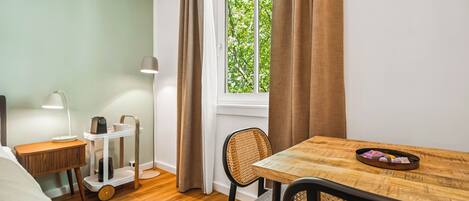 Quarto com mesa de madeira, duas cadeiras, uma mesinha de cabeceira com abajur, janela com cortinas e vista para o verde lá fora.