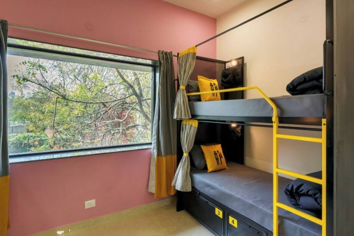 Quarto de dormitório com beliches, janela com cortinas e vista para árvores.