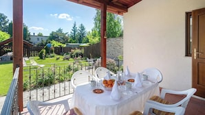 Um alpendre coberto com uma mesa posta para refeições, rodeado por cadeiras brancas, e uma vista para um jardim e edifícios ao fundo.