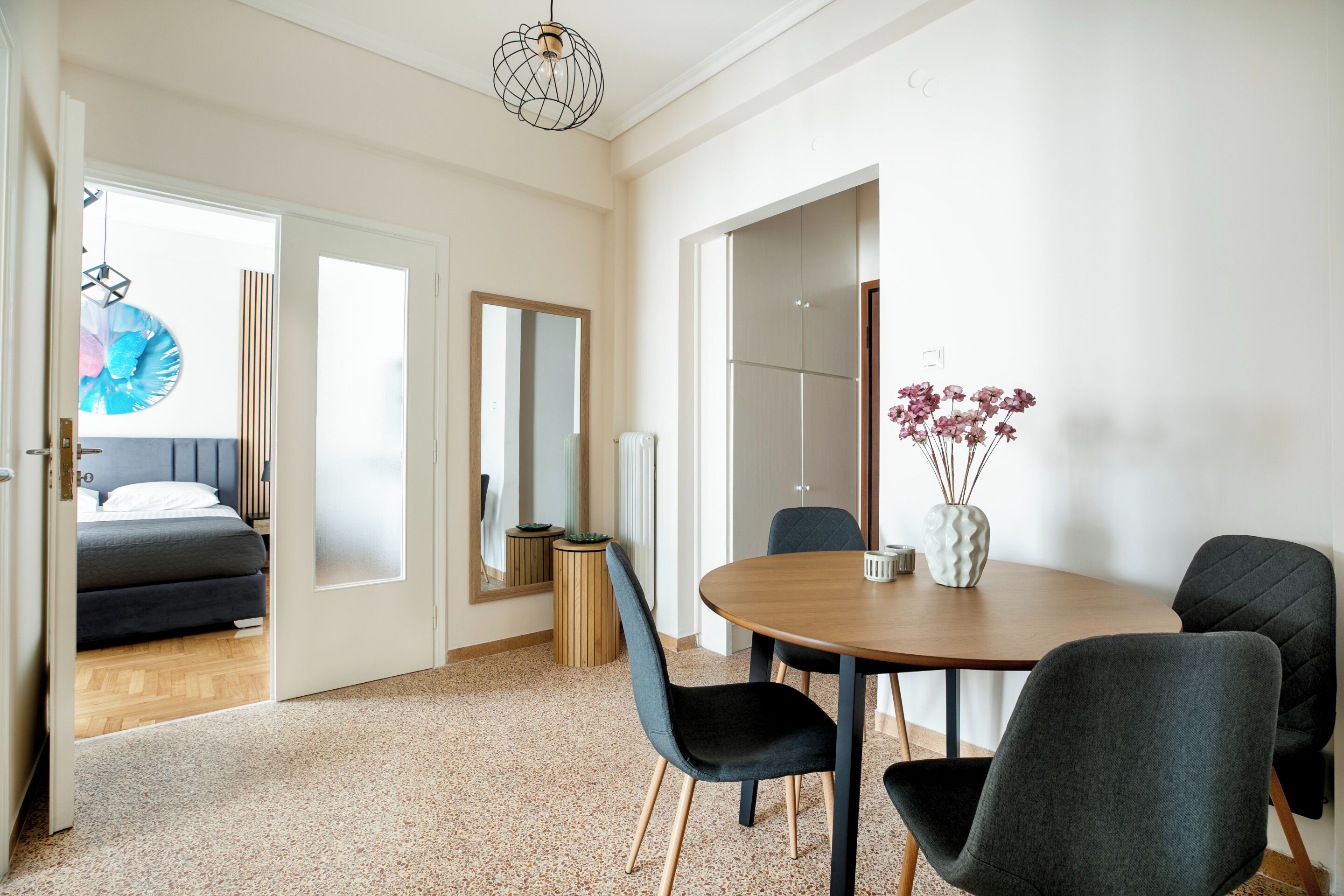 Quarto de hotel moderno com uma mesa de jantar de madeira, quatro cadeiras, um vaso com flores e um espelho grande.