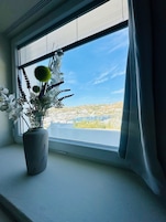 Janela com vista para paisagem costeira, um vaso com flores brancas e um ventilador de teto.
