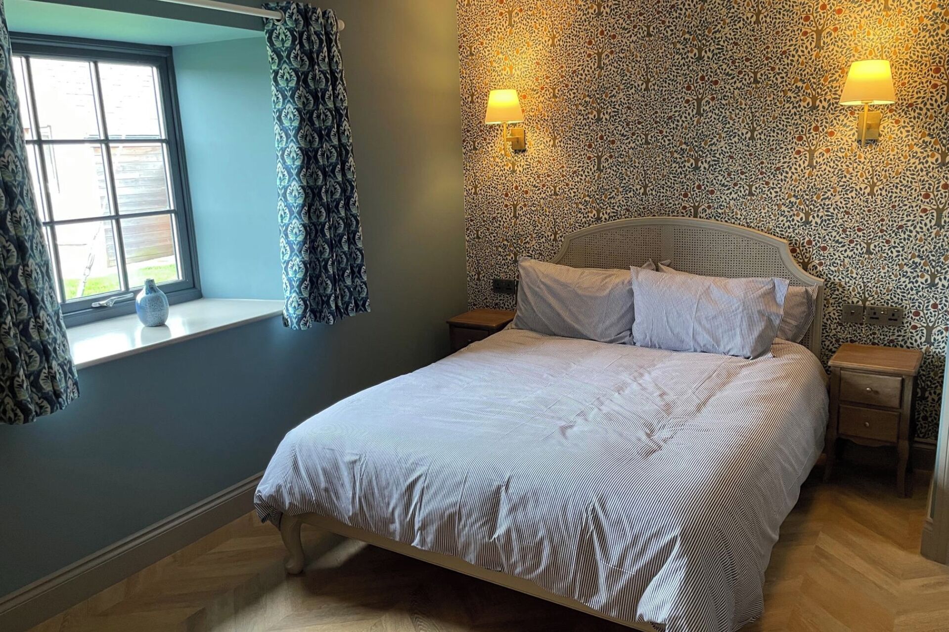 Quarto com cama, duas luminárias de cabeceira, janela com cortinas e parede estampada.