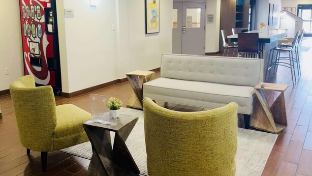 Um saguão de hotel com um sofá branco, duas poltronas verdes, uma mesinha com uma planta e uma máquina de vendas.