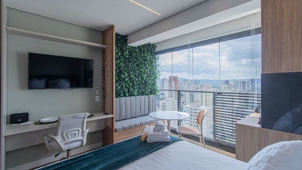 Quarto de hotel moderno com uma janela ampla, uma cama, uma escrivaninha, uma cadeira e uma televisão.