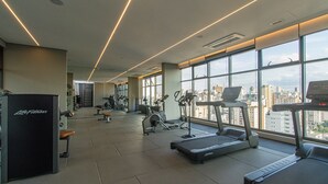 Uma academia moderna com diversos equipamentos de exercício, amplas janelas com vista para a cidade e um layout espaçoso e bem cuidado.