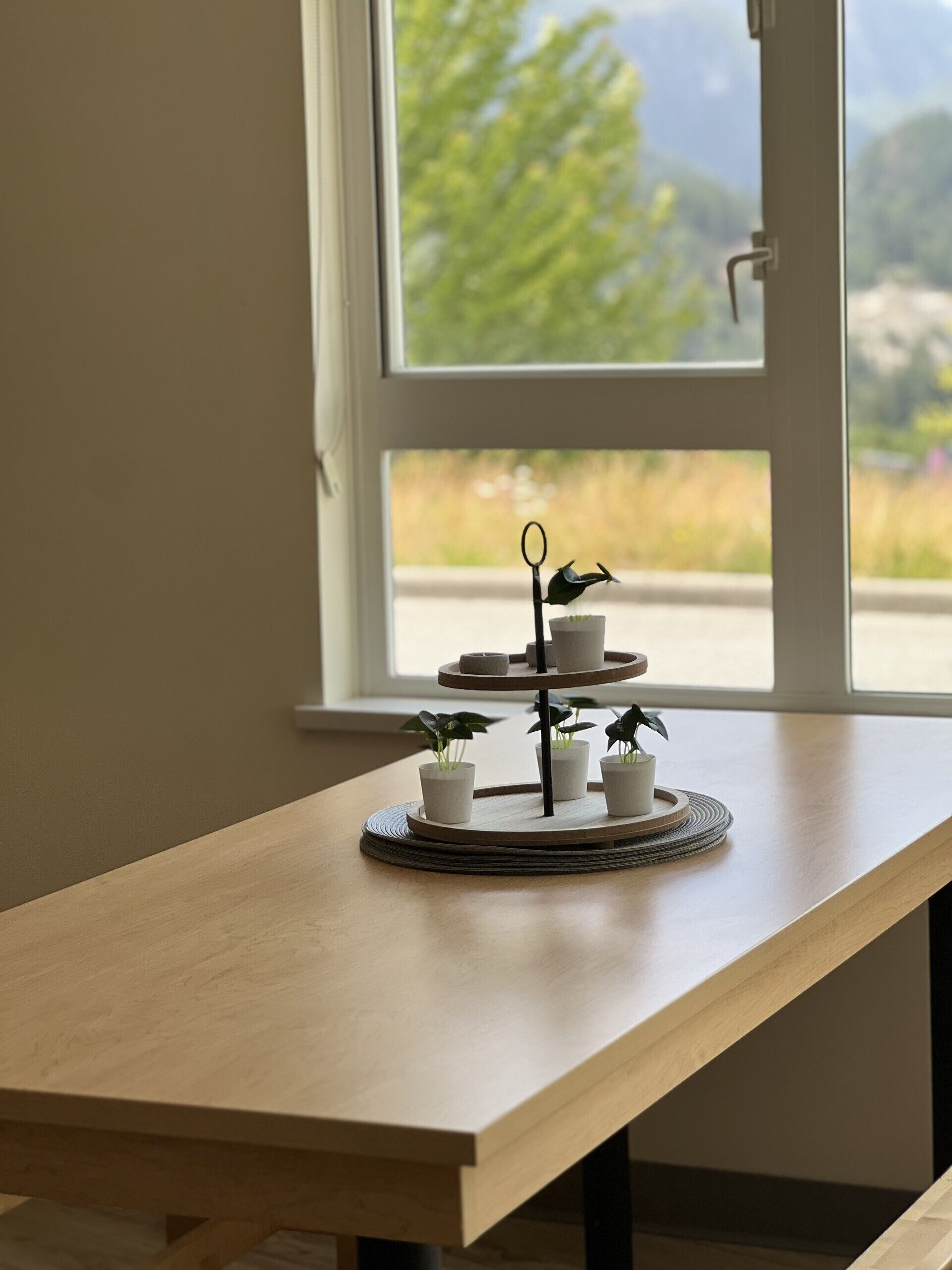 Uma mesa de madeira com um suporte escalonado para pequenos vasos de plantas, colocada perto de uma janela com vista para vegetação e montanhas.