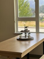 Uma mesa de madeira com um suporte escalonado para pequenos vasos de plantas, colocada perto de uma janela com vista para vegetação e montanhas.