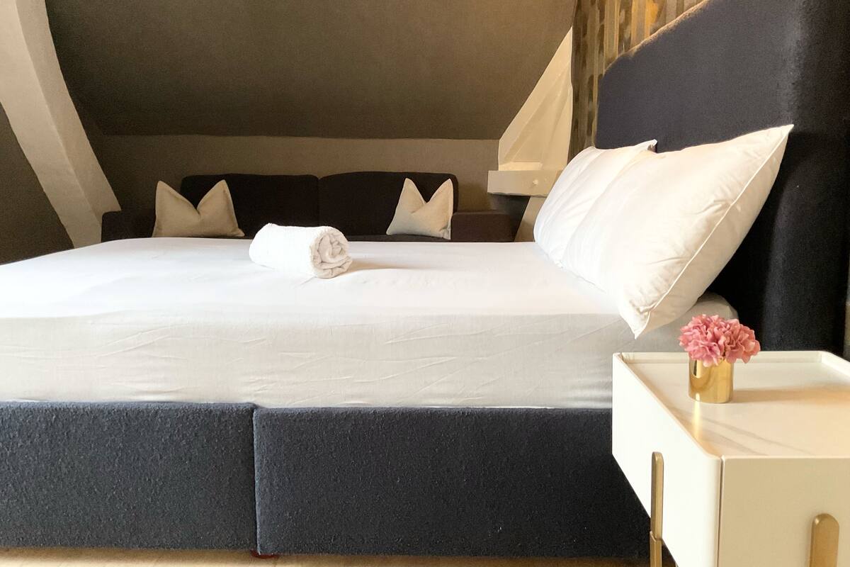 Cama bem arrumada com lençóis brancos e uma toalha enrolada, cabeceira estofada preta e mesa de cabeceira com um vaso de flores cor-de-rosa.