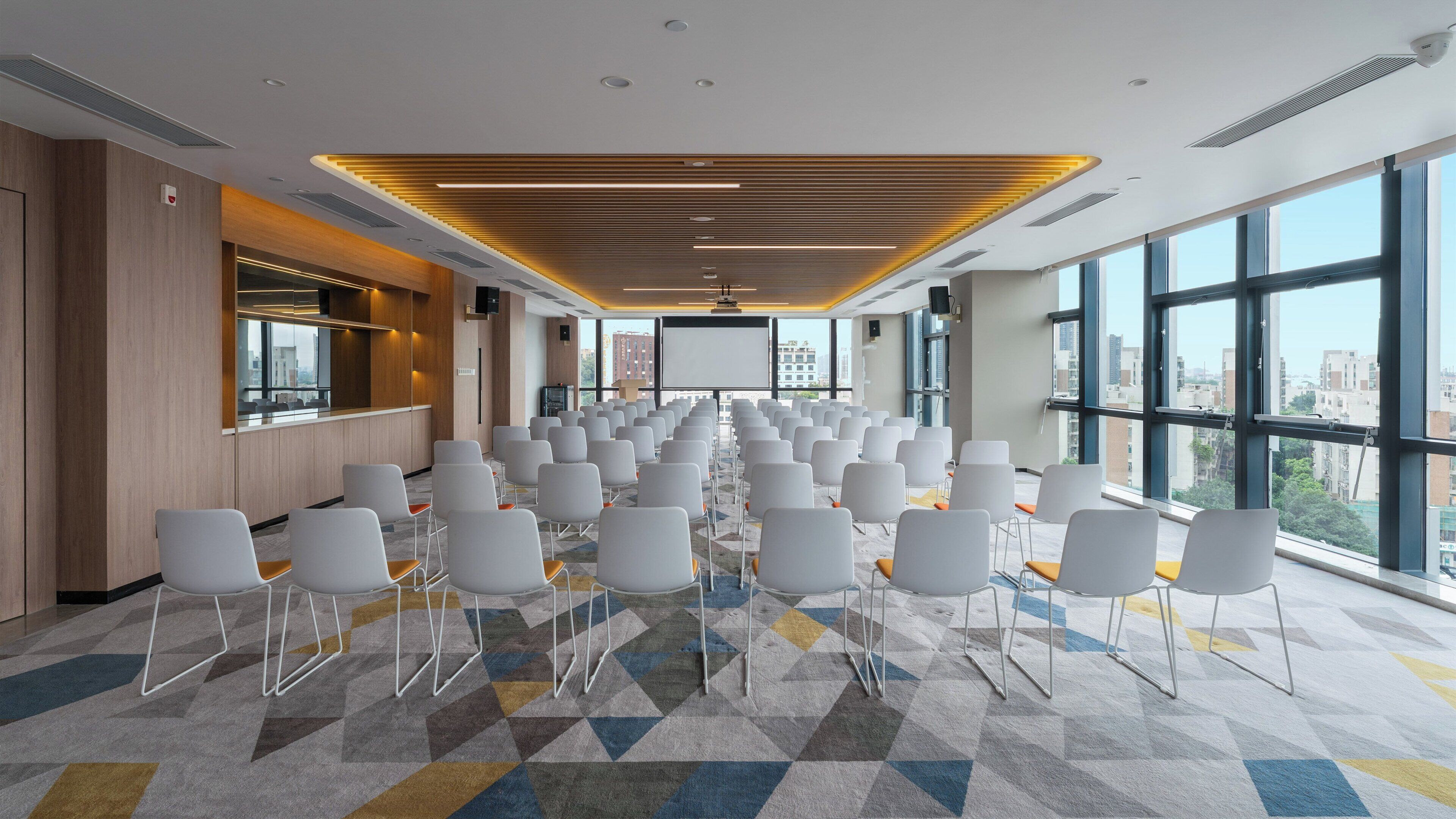 Sala de conferências com fileiras de cadeiras, uma tela grande e vista para a cidade através de amplas janelas.
